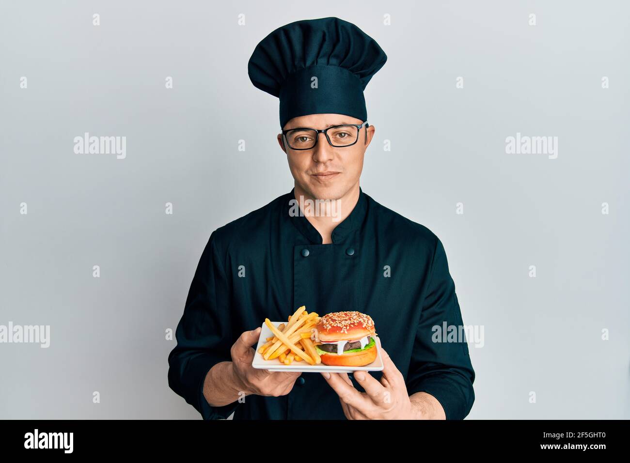 Handsome young man chef holding burger with fries relaxed with serious ...