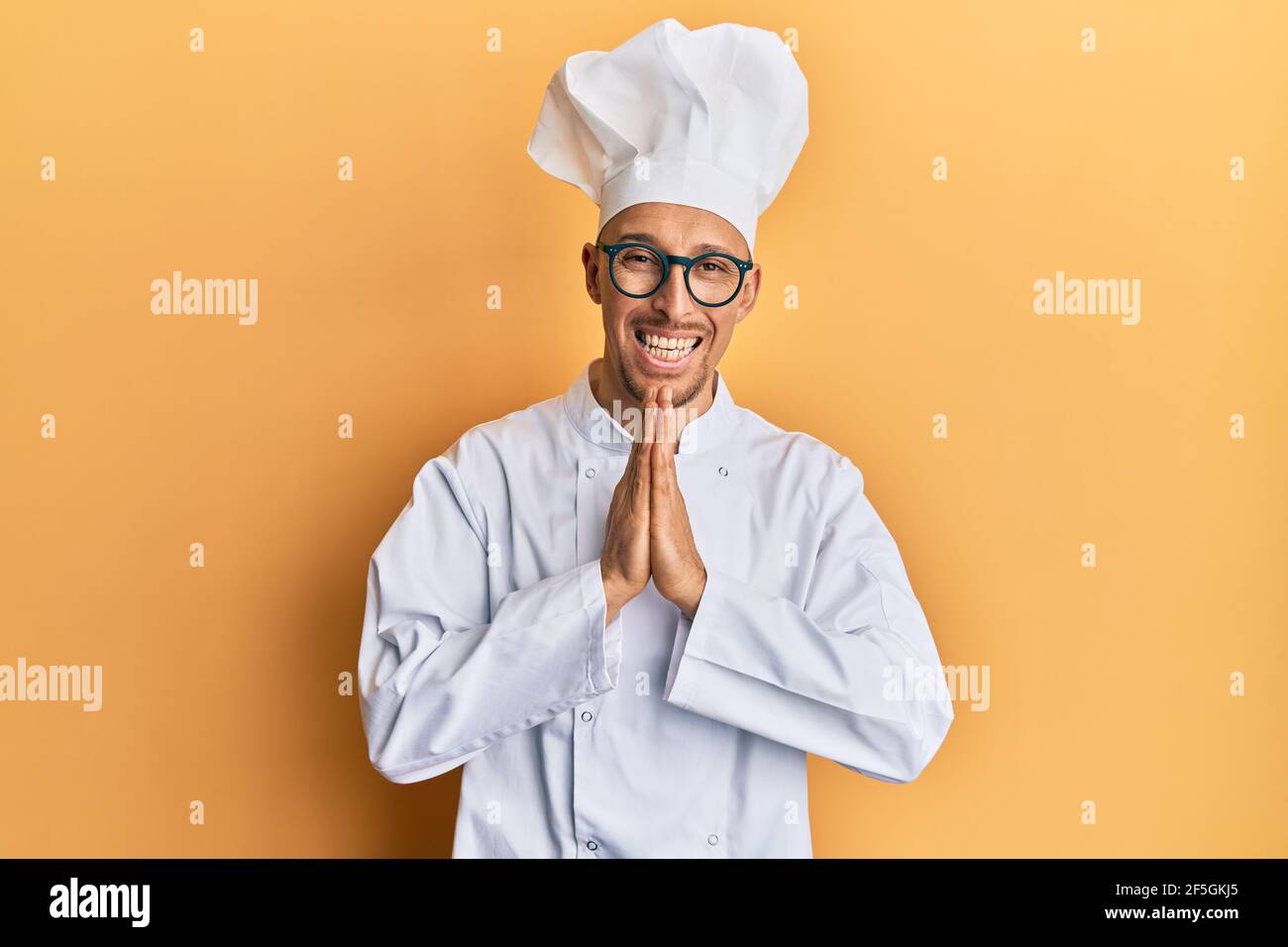 Bald man with beard wearing professional cook uniform praying with ...