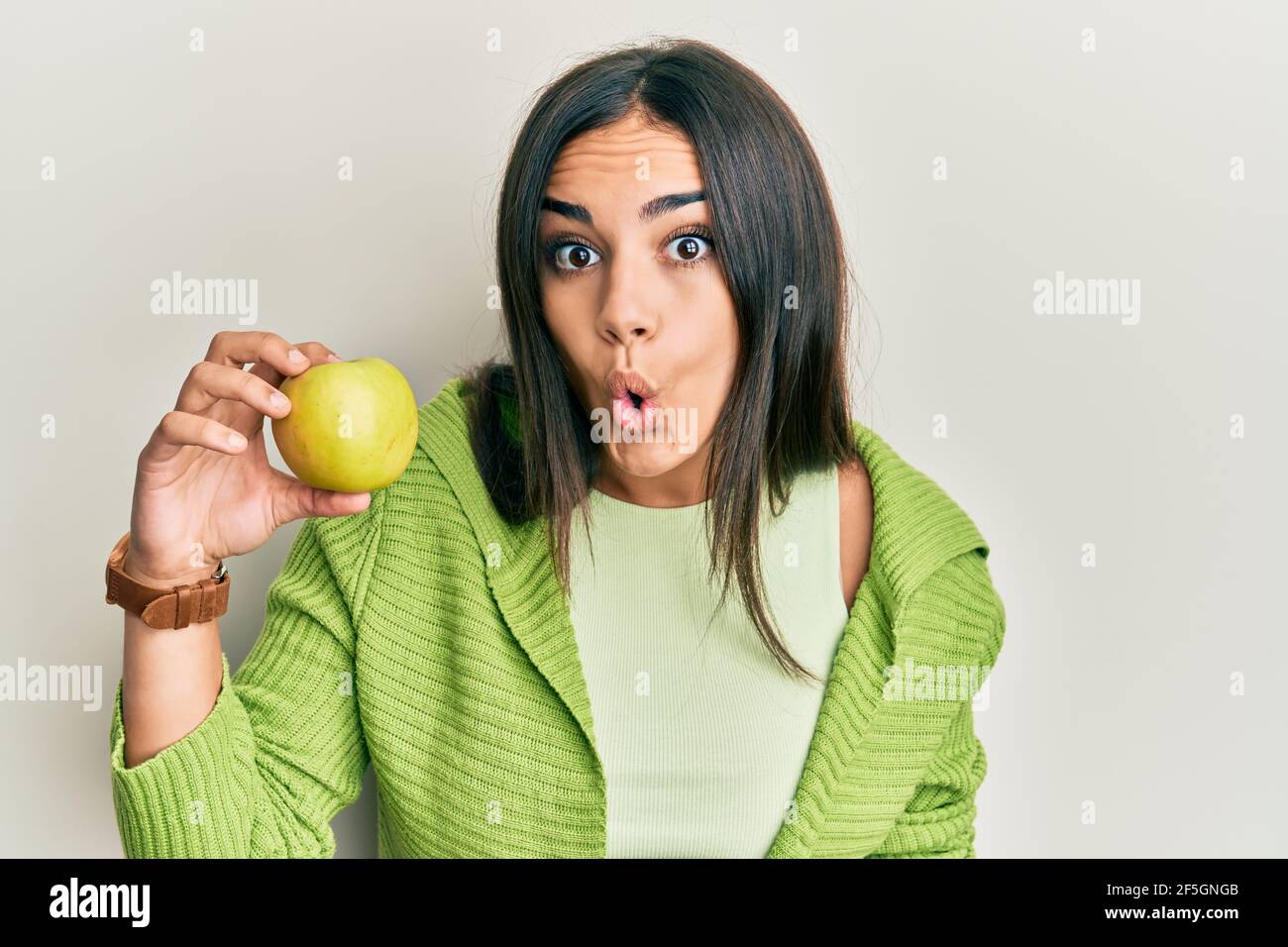 Young brunette woman holding green apple scared and amazed with open ...