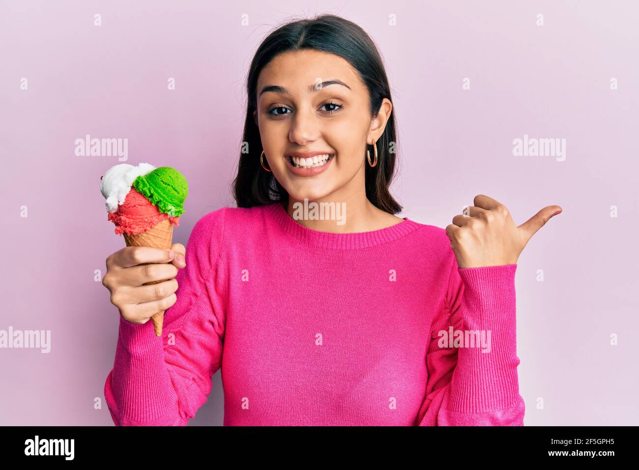 Young hispanic woman holding ice cream pointing thumb up to the side ...