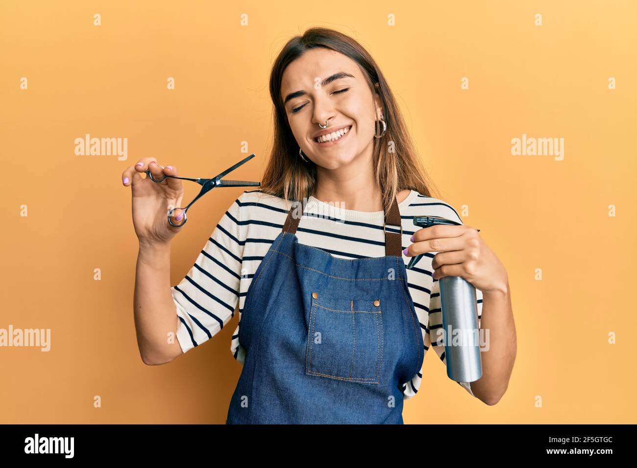 Young hispanic woman wearing hairdresser apron and holding scissors smiling and laughing hard ...