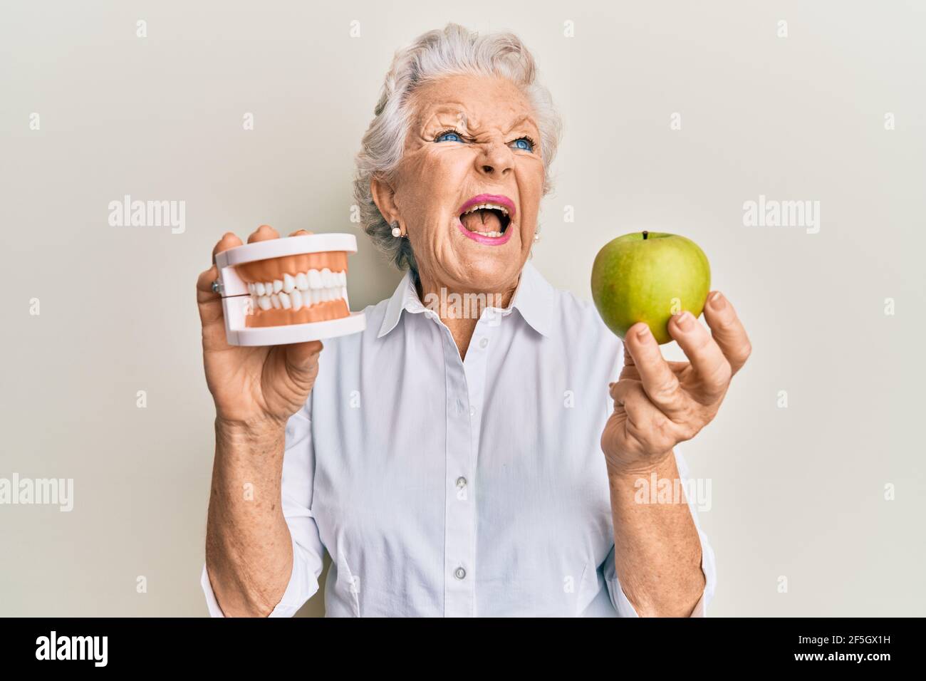 Senior grey-haired woman holding green apple and denture teeth angry ...
