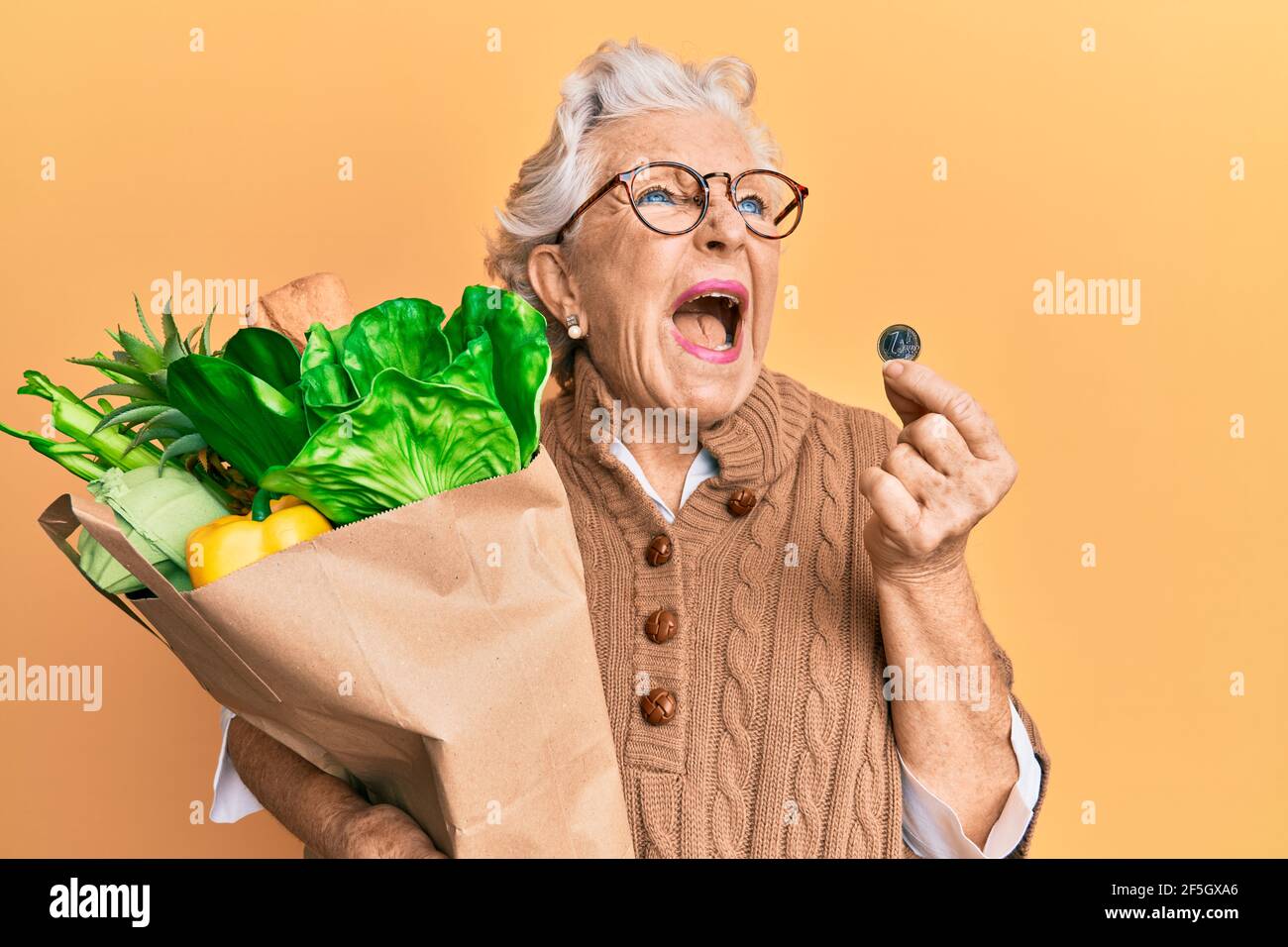 Senior grey-haired woman holding groceries and 1 euro coin angry and ...
