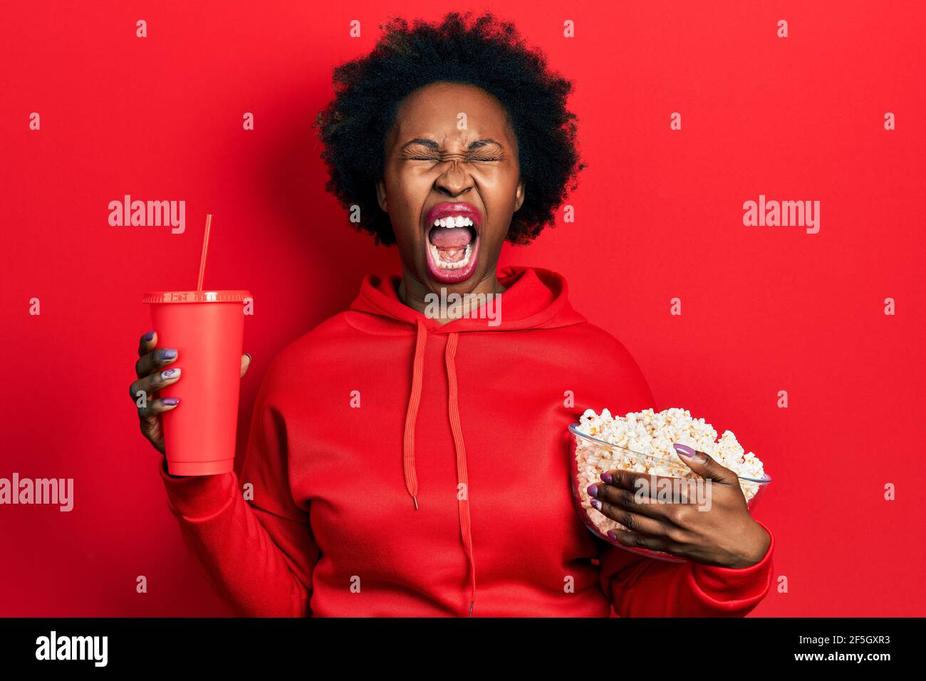 Young african american woman eating popcorn and drinking soda angry and ...