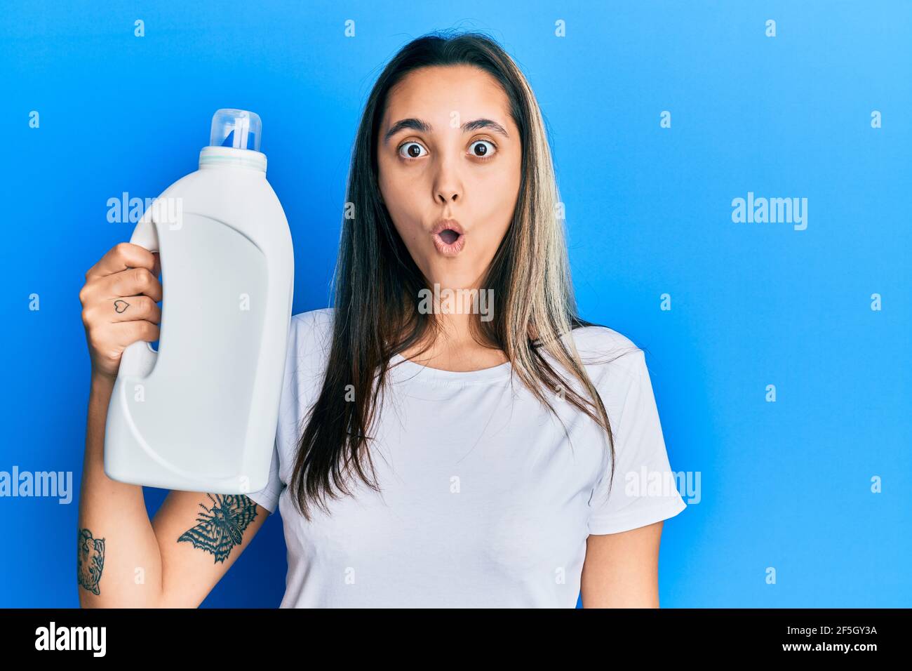 Young hispanic woman holding detergent bottle scared and amazed with open mouth for surprise ...