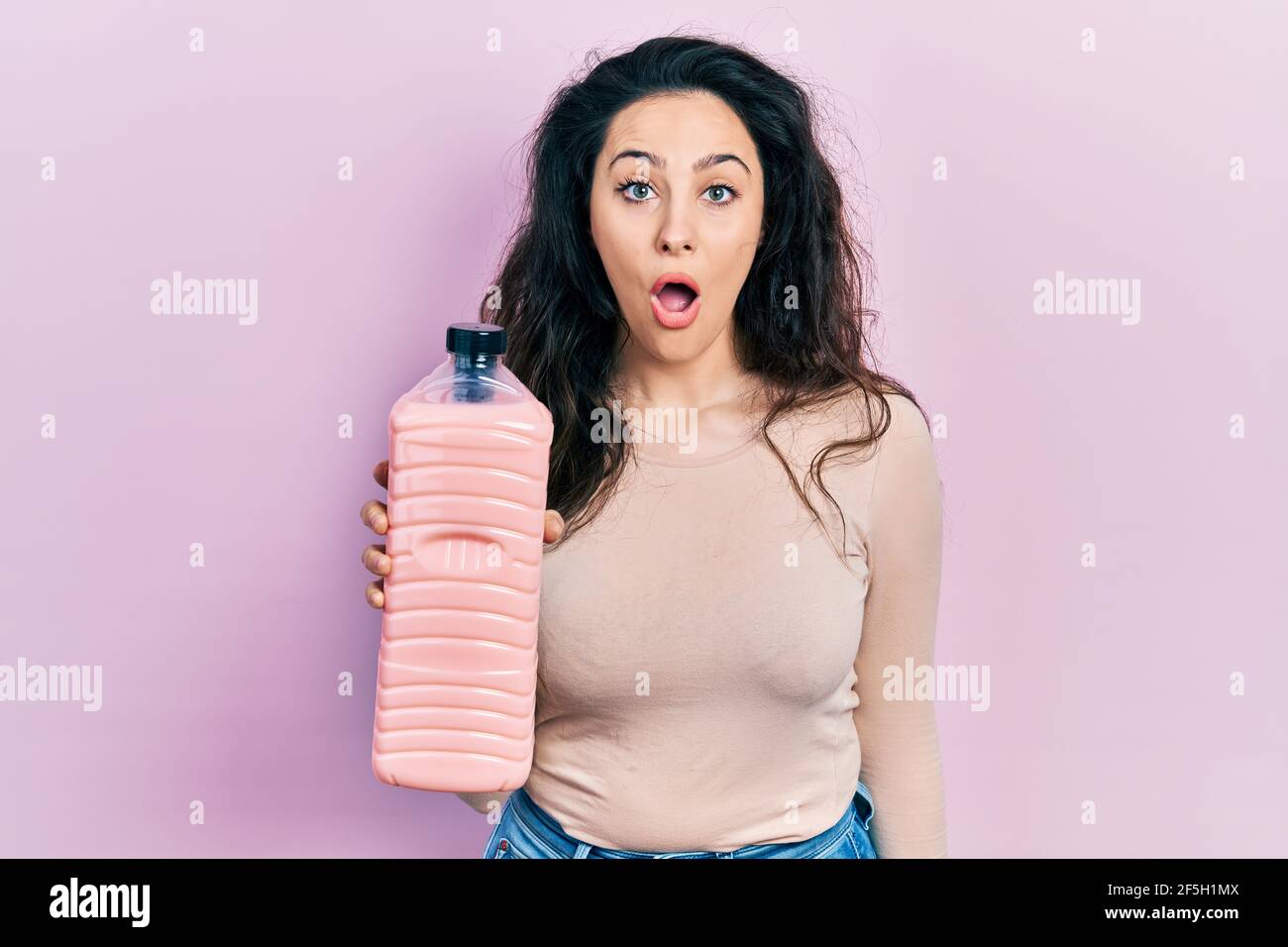Young hispanic woman holding detergent bottle scared and amazed with open mouth for surprise ...