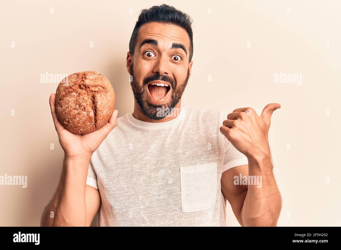 Young hispanic man holding bread pointing thumb up to the side smiling ...