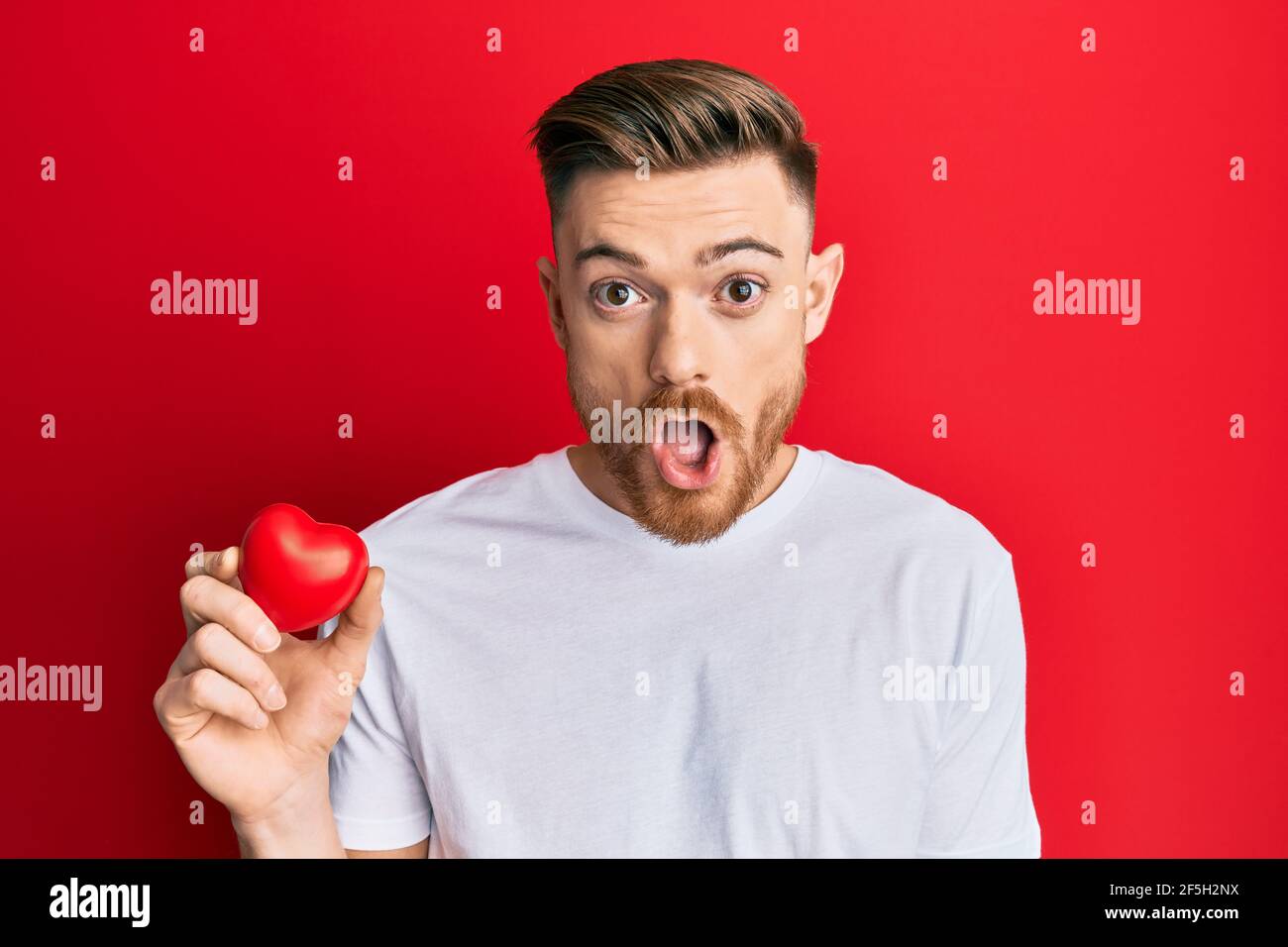 Young redhead man holding heart scared and amazed with open mouth for ...