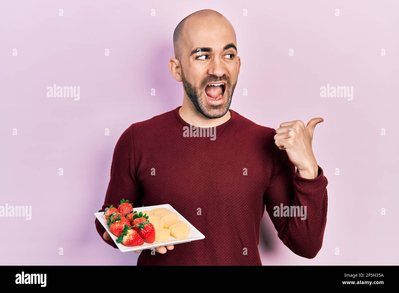 Young bald man eating fresh and healthy fruits pointing thumb up to the ...