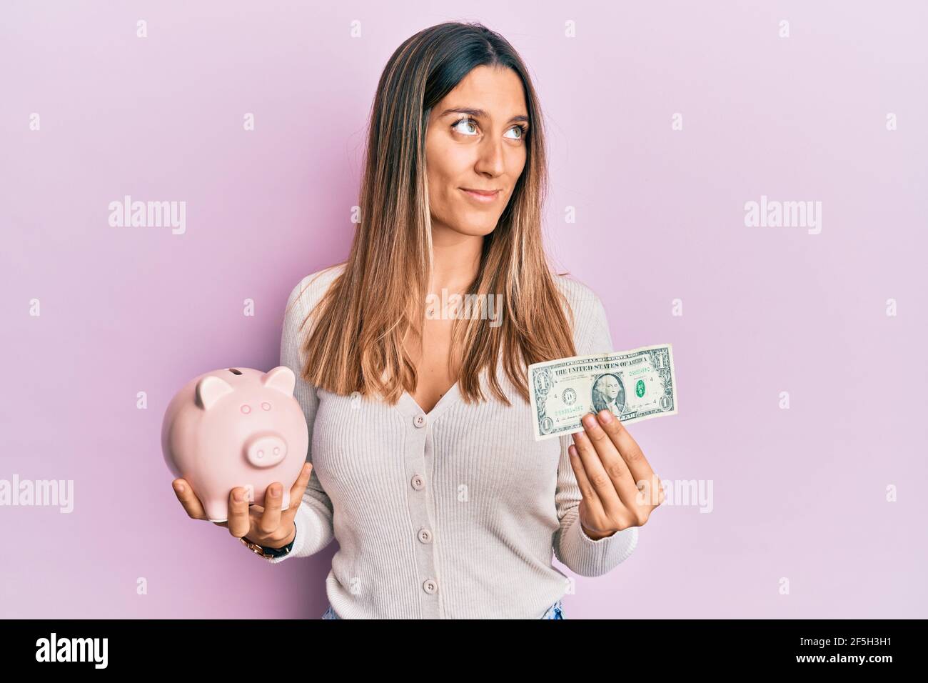 Brunette young woman holding one dollar banknote and piggy bank smiling ...