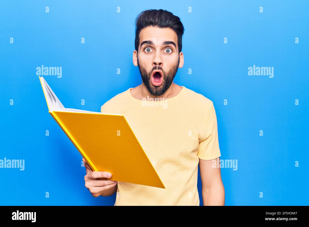 Young handsome man with beard reading book scared and amazed with open ...