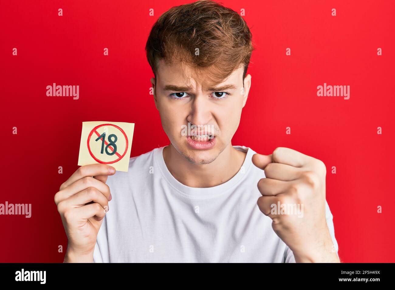 Young caucasian man holding under 18 prohibition sticker annoyed and ...