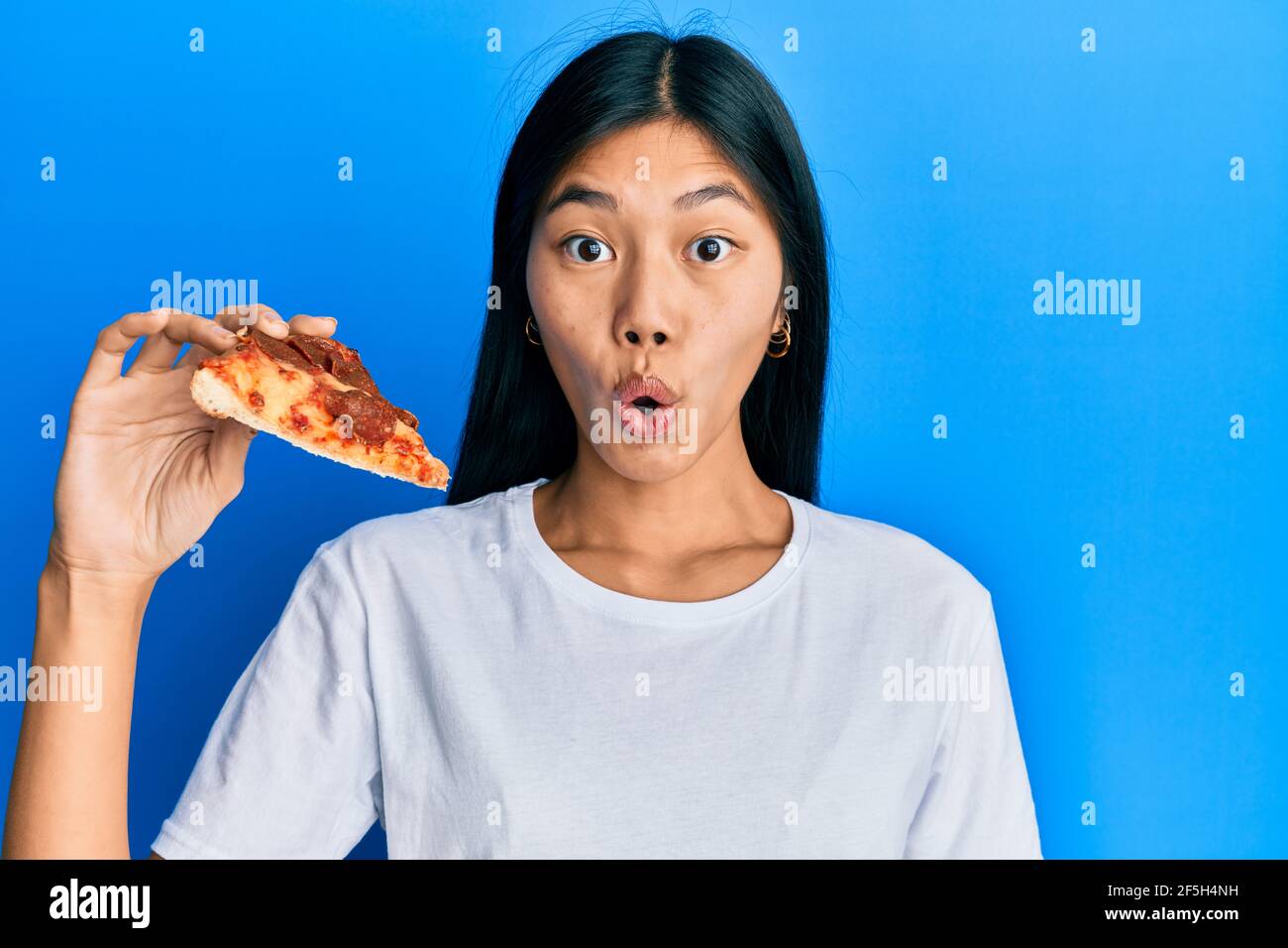 Young chinese woman eating tasty pepperoni pizza scared and amazed with ...