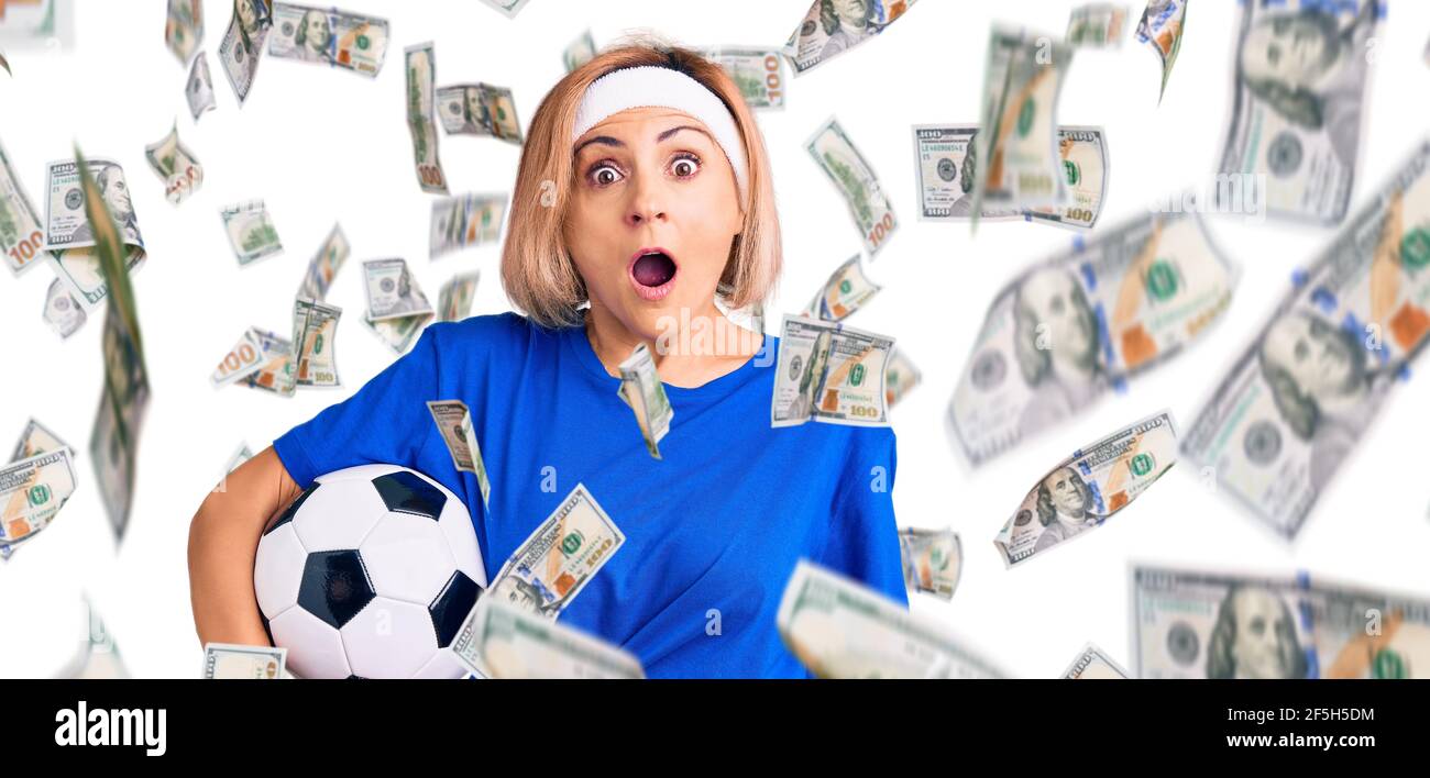 Young blonde woman holding soccer ball scared and amazed with open ...