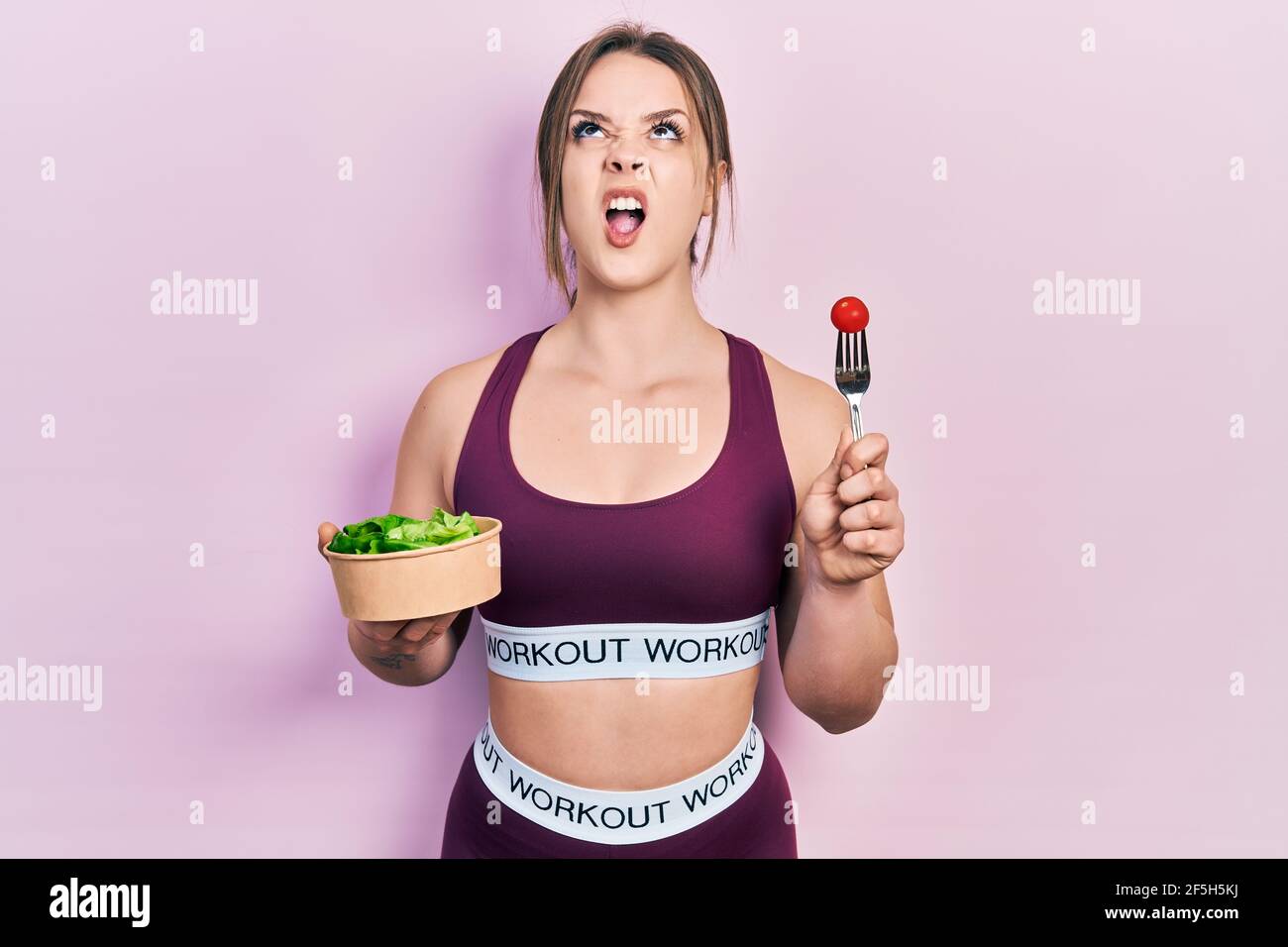 Young hispanic girl wearing sportswear eating salad angry and mad ...