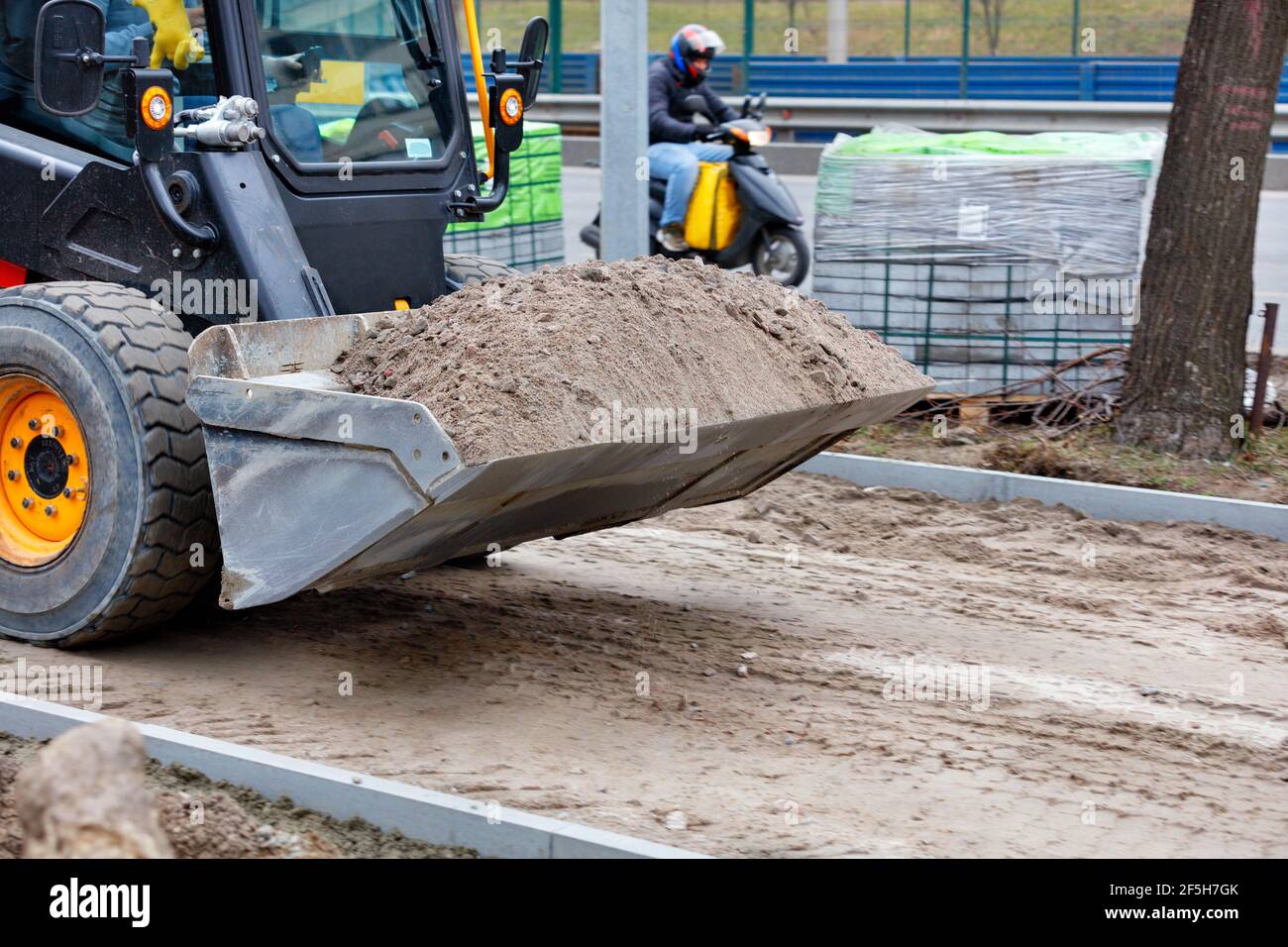 A small grader moves along a sidewalk under construction and carries a ...