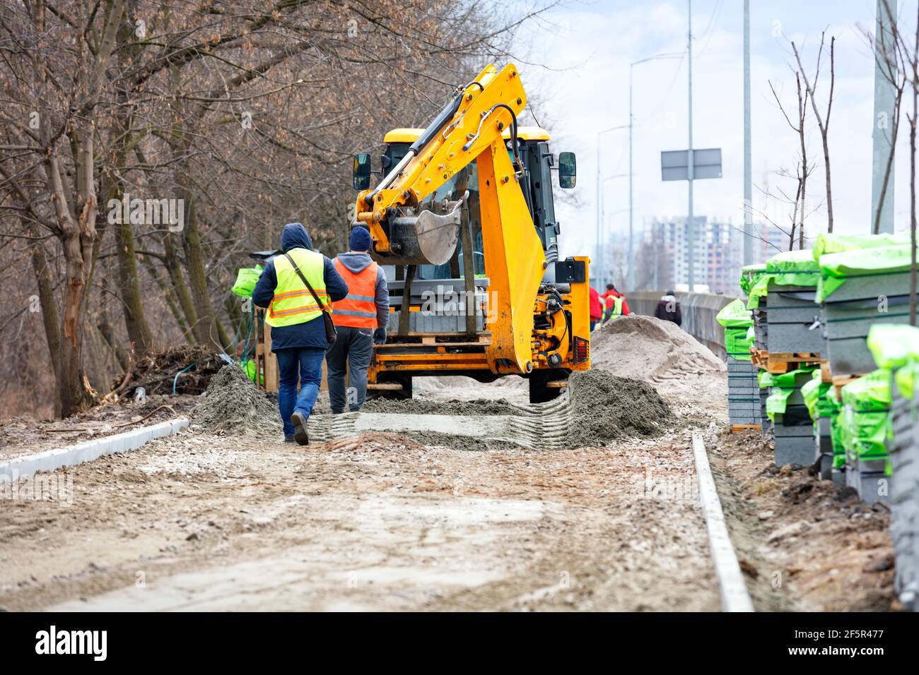 An orange road excavator and a group of workers prepare a sandy base ...