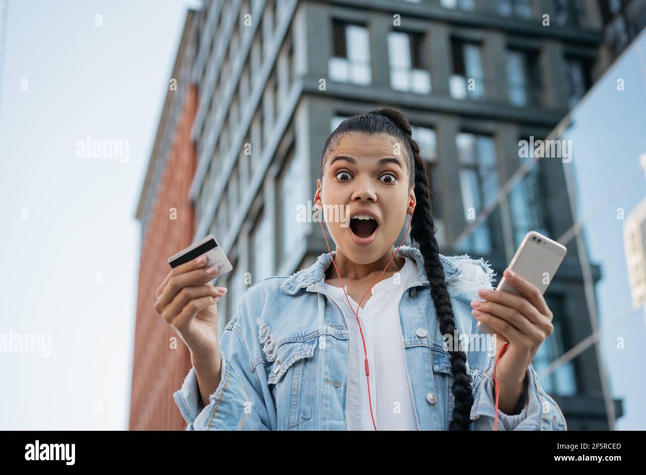 African American woman with emotional face and open mouth holding credit card, using smartphone ...
