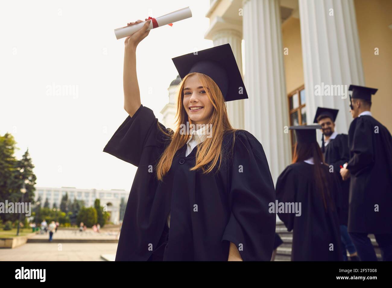 Happy smiling graduate girl dressed in black gown raises up her hand holding a diploma Stock ...