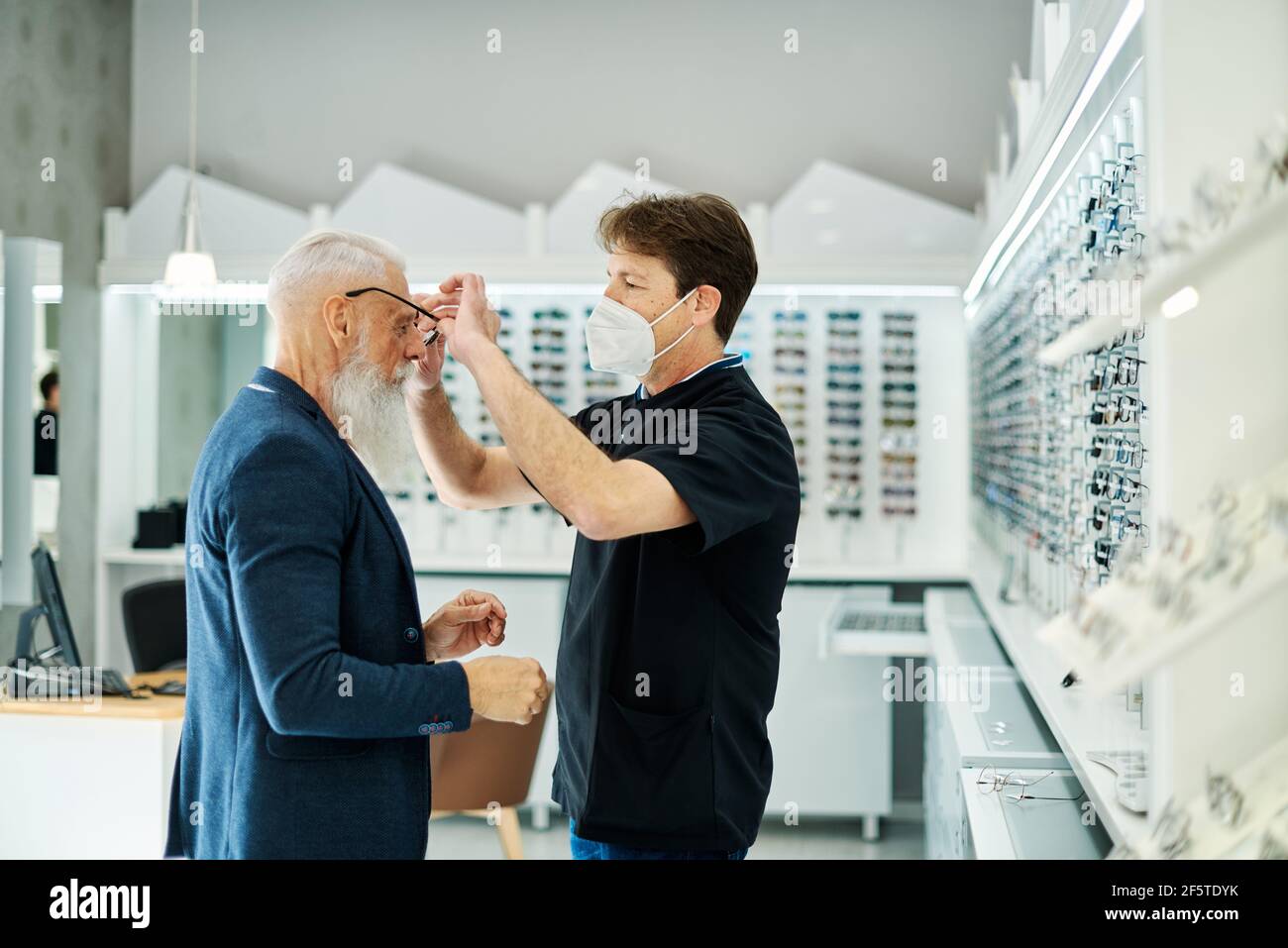Side view of professional optician in mask helping senior male putting ...