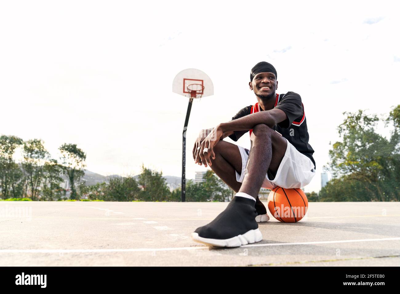 Low angle of smiling black male streetball player sitting on ball on ...