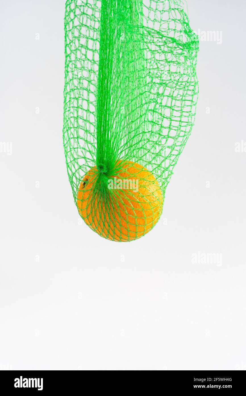 A single orange hanging in a green net bag on white background with ...