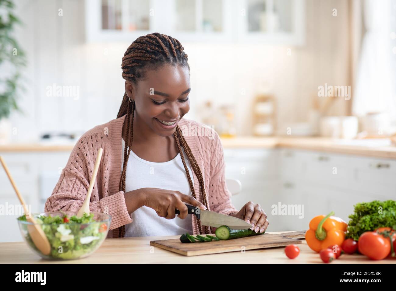 Happy young black woman cooking fresh vegetable salad in kitchen ...