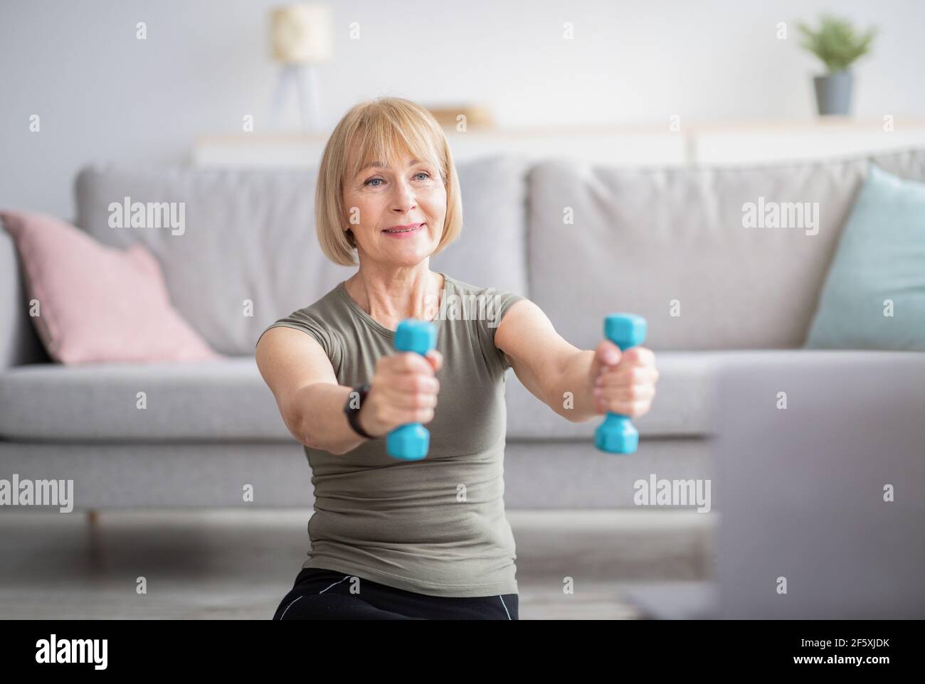 Positive mature woman training with dumbbells near laptop at home ...