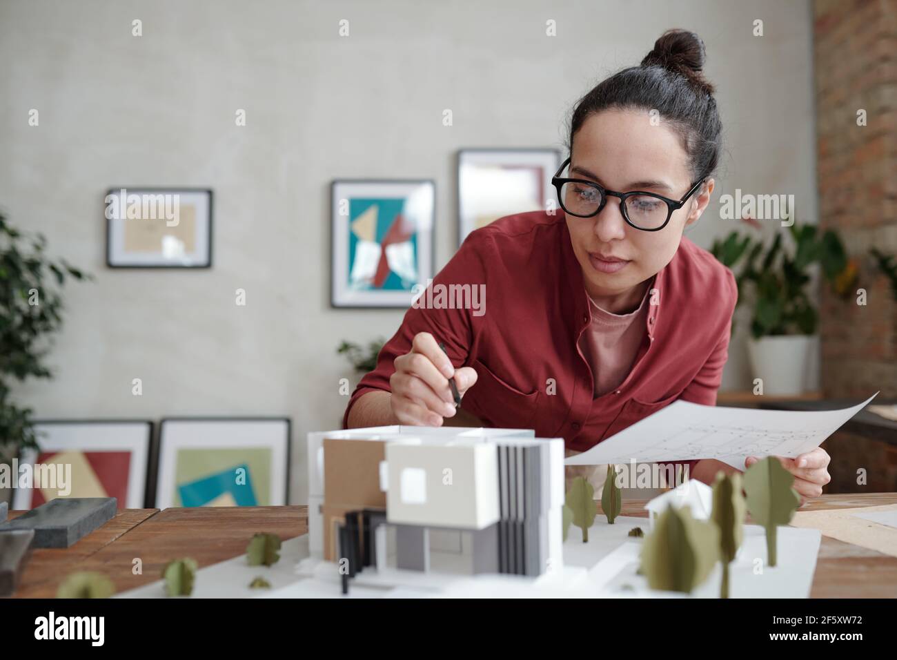 Young elegant female architect with paper and pen bending over model of ...