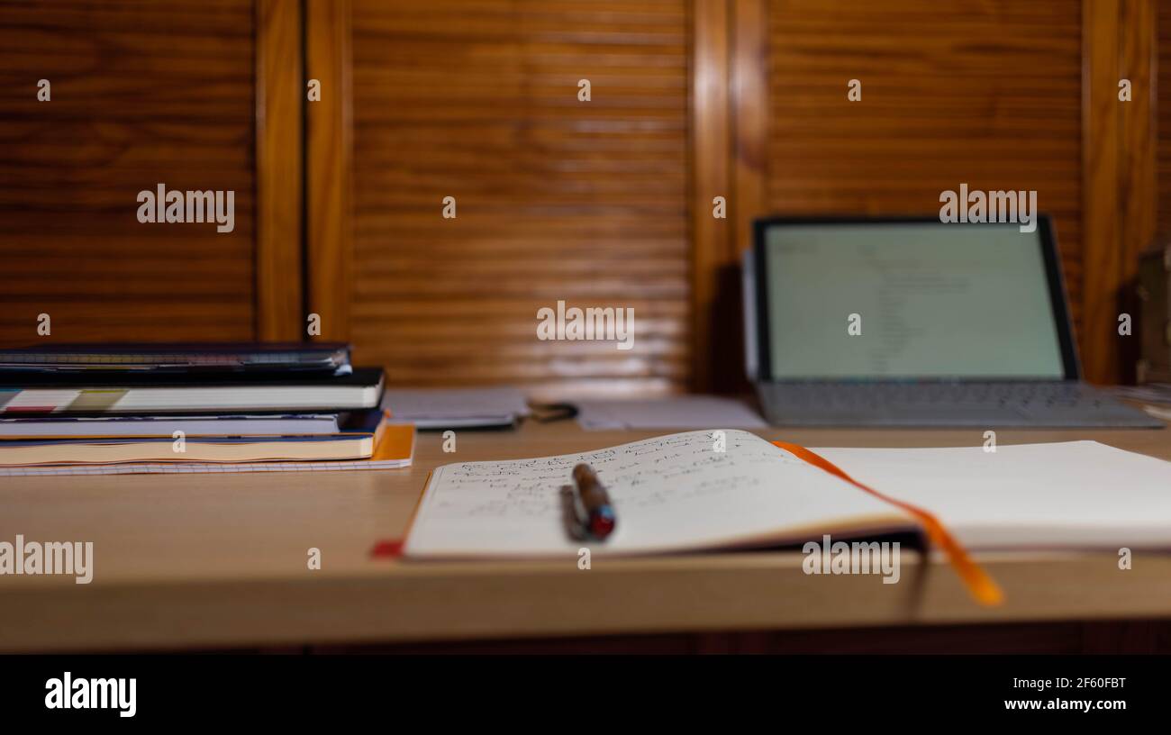 a stack of paper notebooks piled on a wooden desk. a screen in the ...