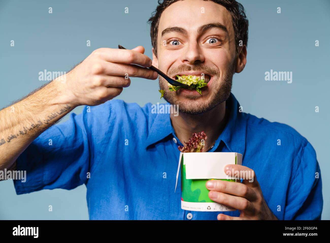 Excited handsome hungry guy eating salad takeaway isolated over blue ...