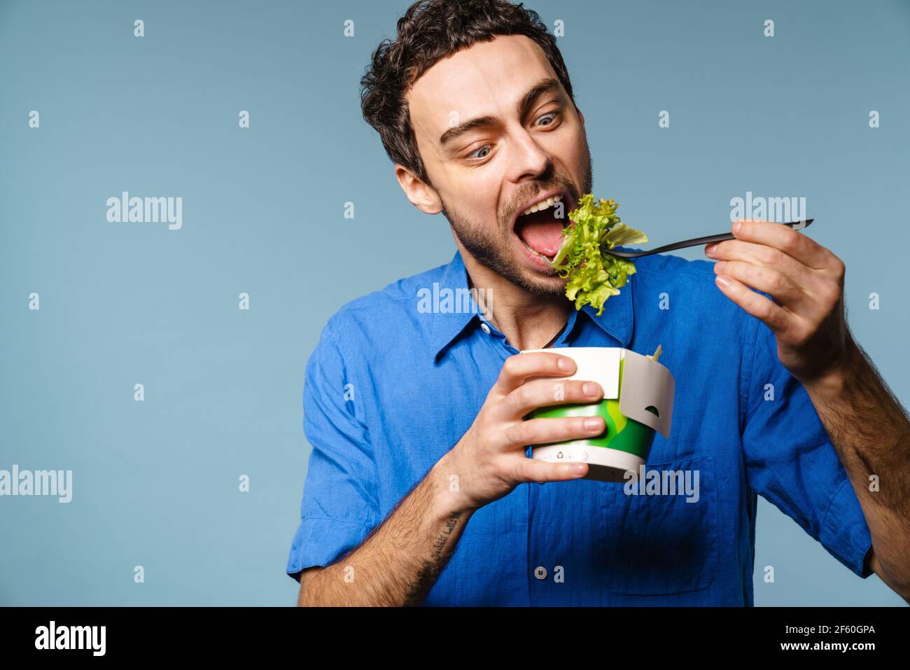 Excited handsome hungry guy eating salad takeaway isolated over blue ...