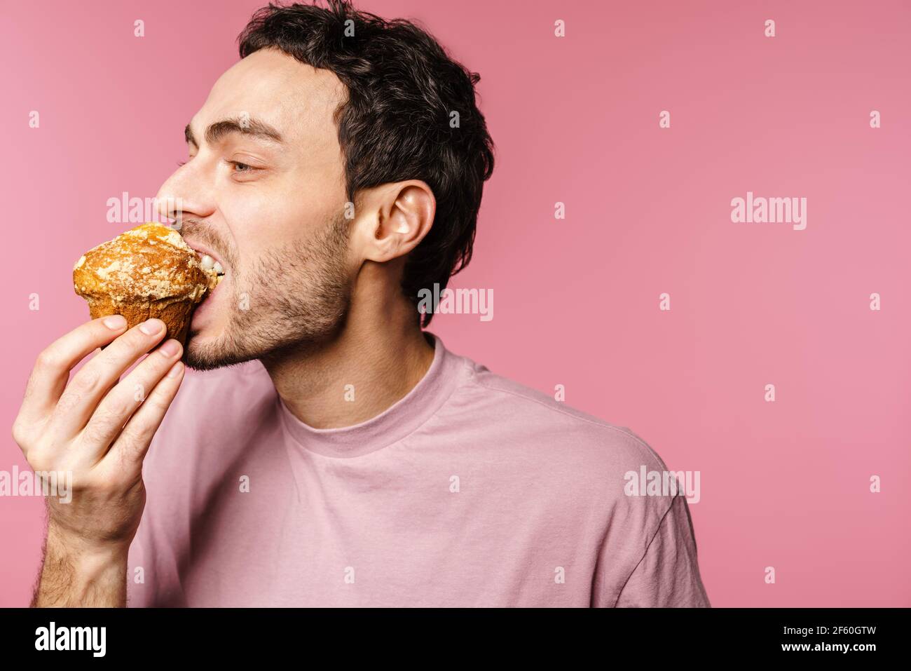 Happy handsome guy eating muffin with eyes closed isolated over pink ...