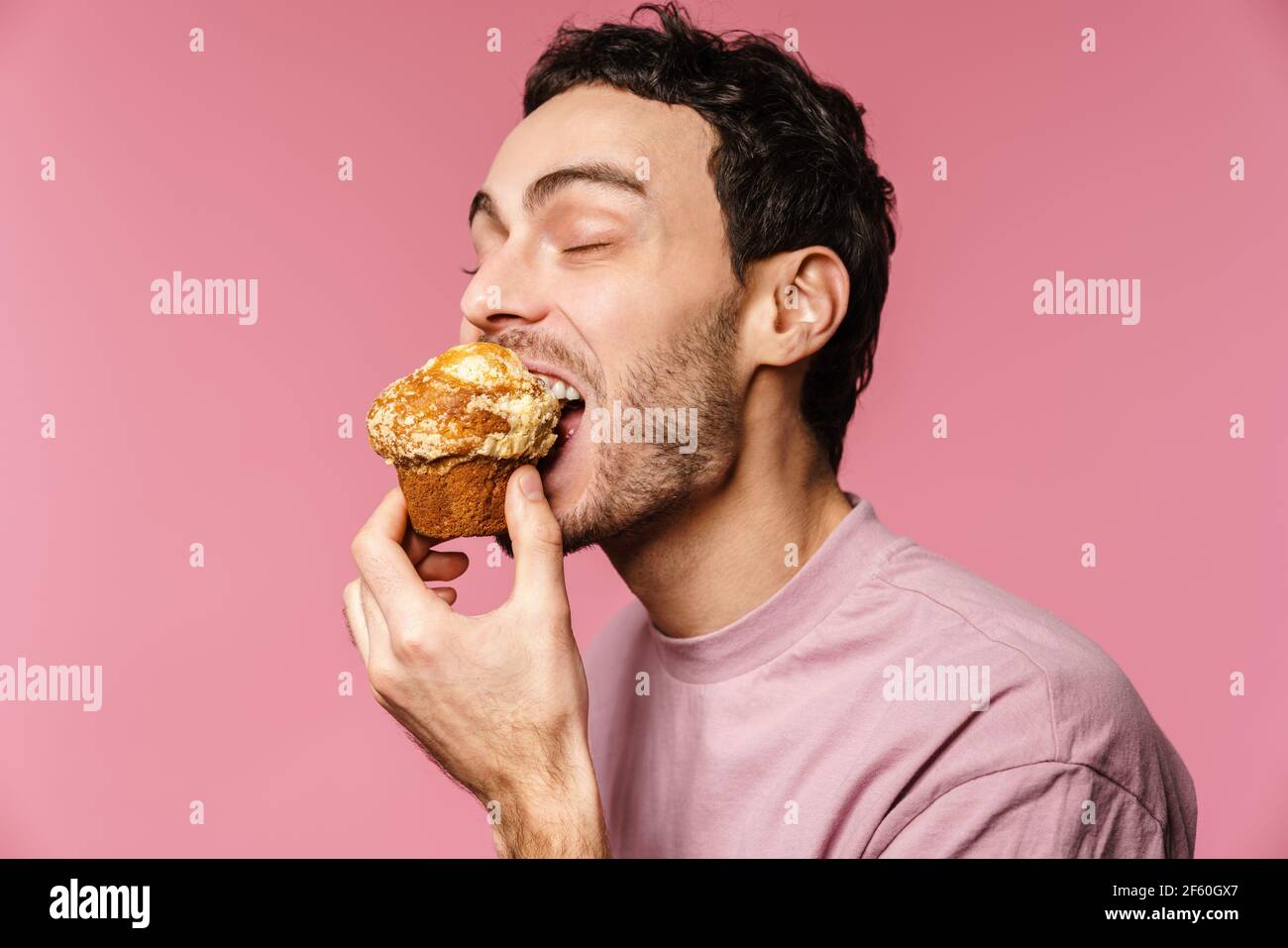 Happy handsome guy eating muffin with eyes closed isolated over pink ...
