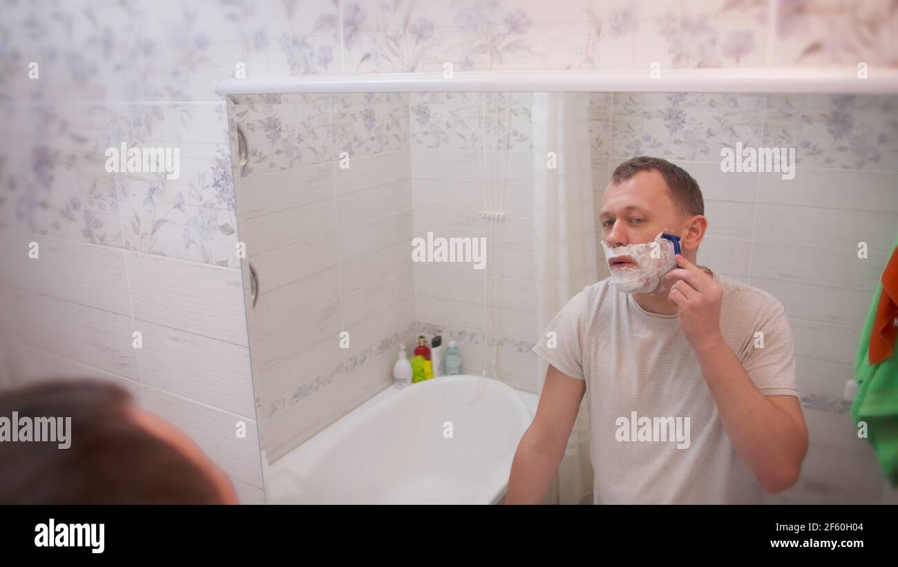 Young attractive man shaving while standing in the bathroom in front of ...