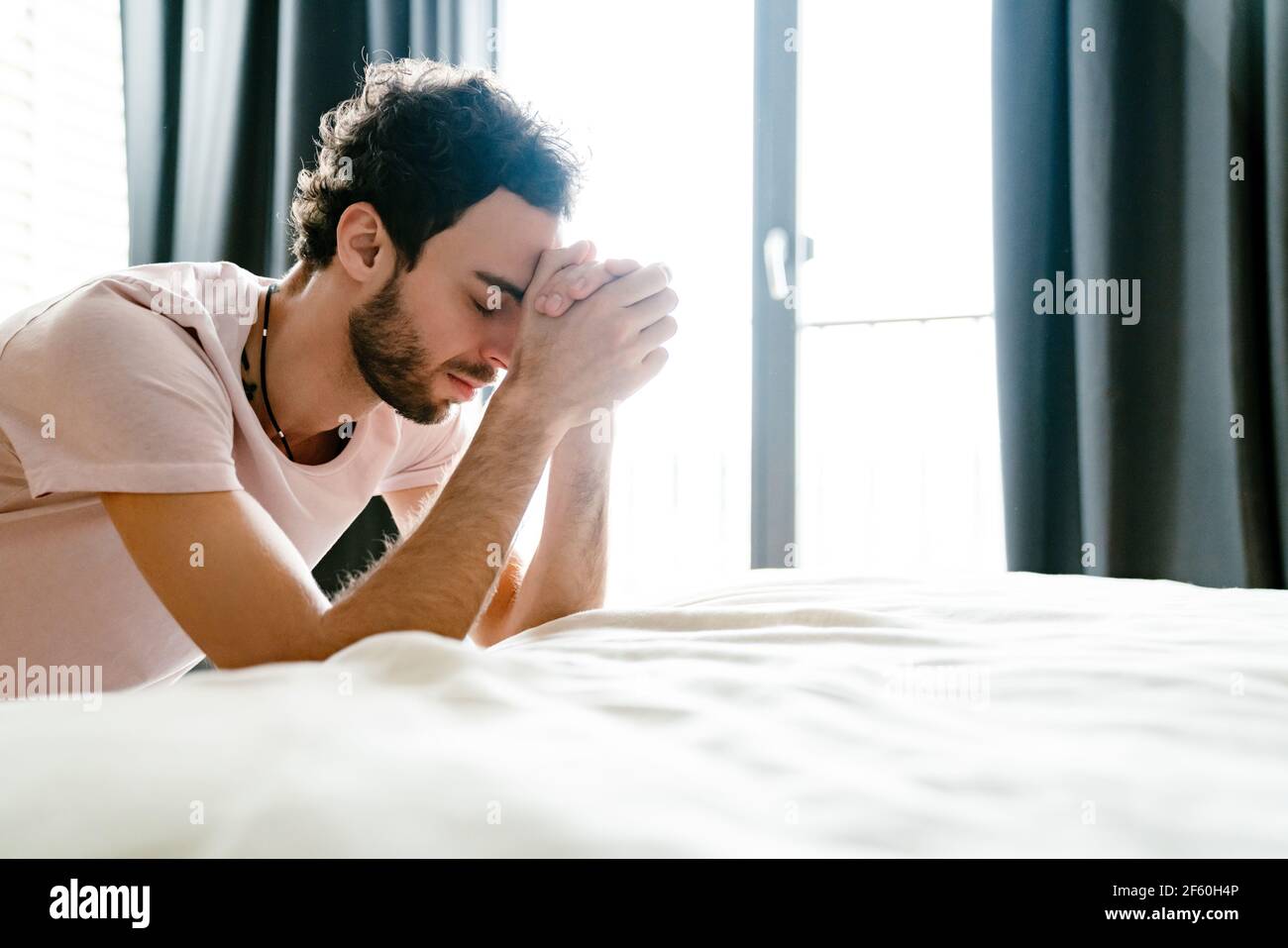 Focused unshaven man praying while holding hands together in bedroom at ...