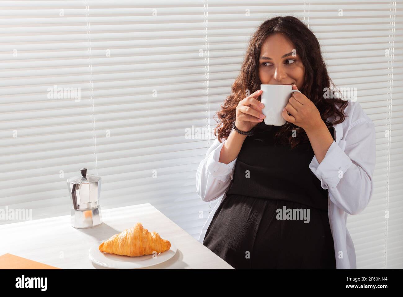 Joyful pregnant young beautiful woman looks through blinds during her ...