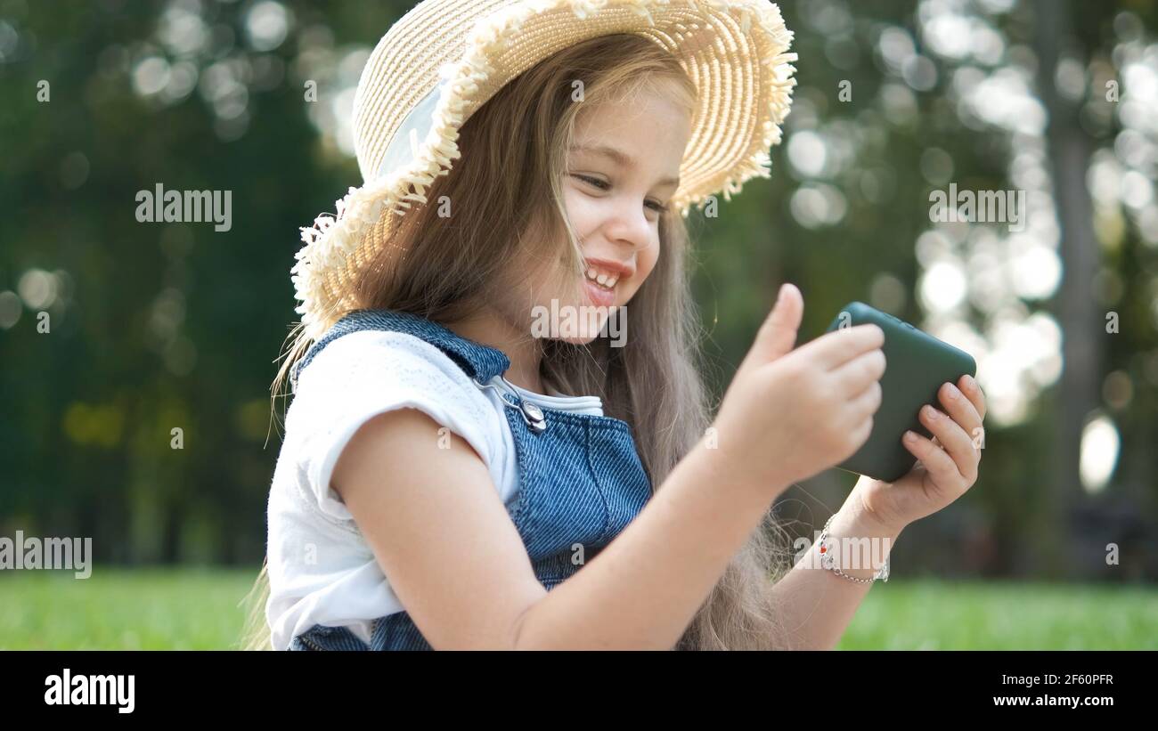 Happy smiling child girl looking in mobile phone outdoors in summer ...