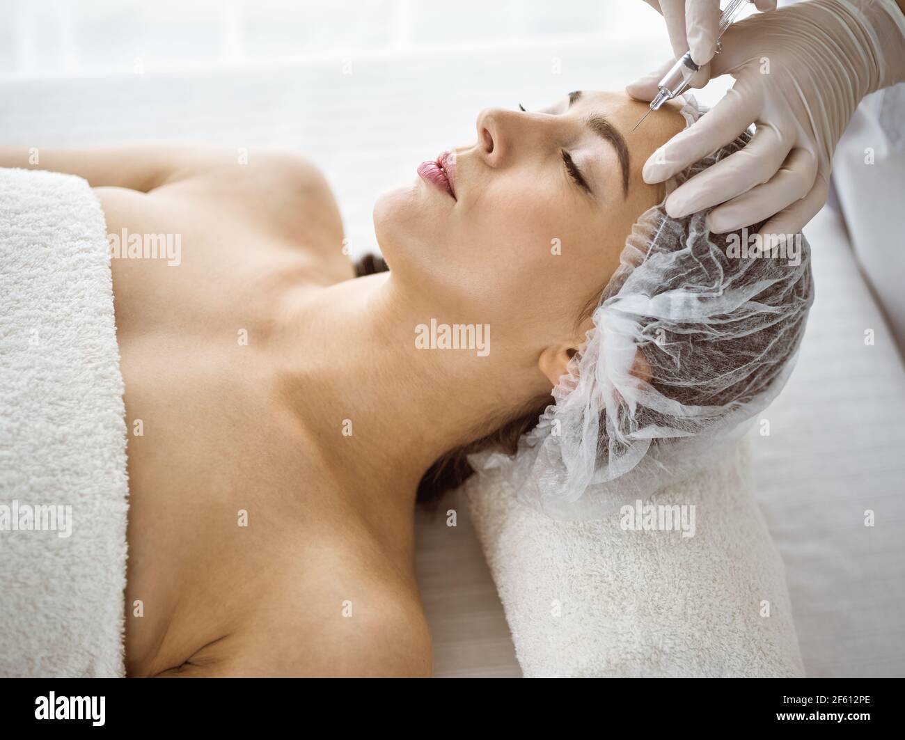 Beautician doing beauty procedure with syringe to face of young brunette woman in sunny clinic ...