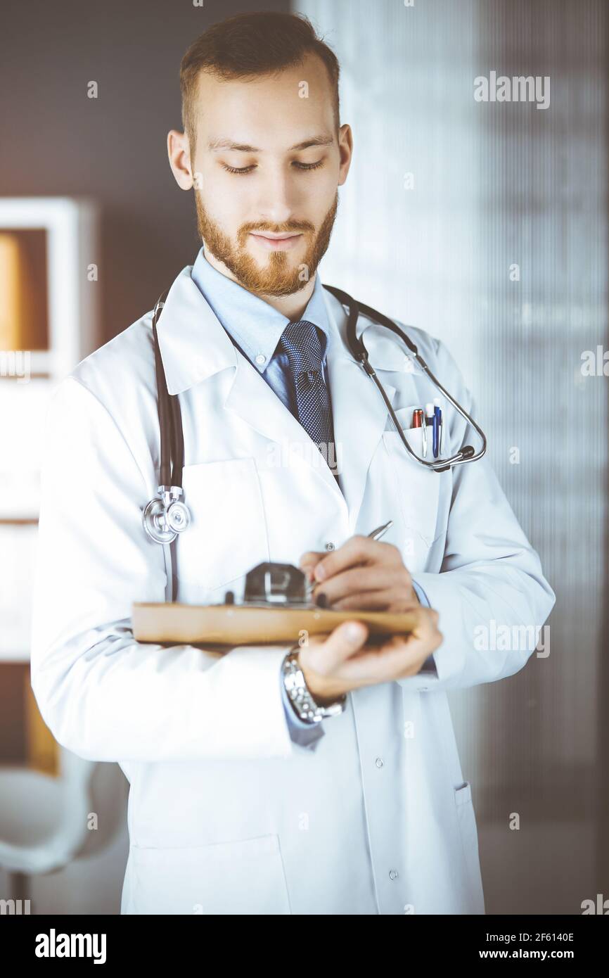 Friendly red-bearded doctor standing and writing with clipboard in ...