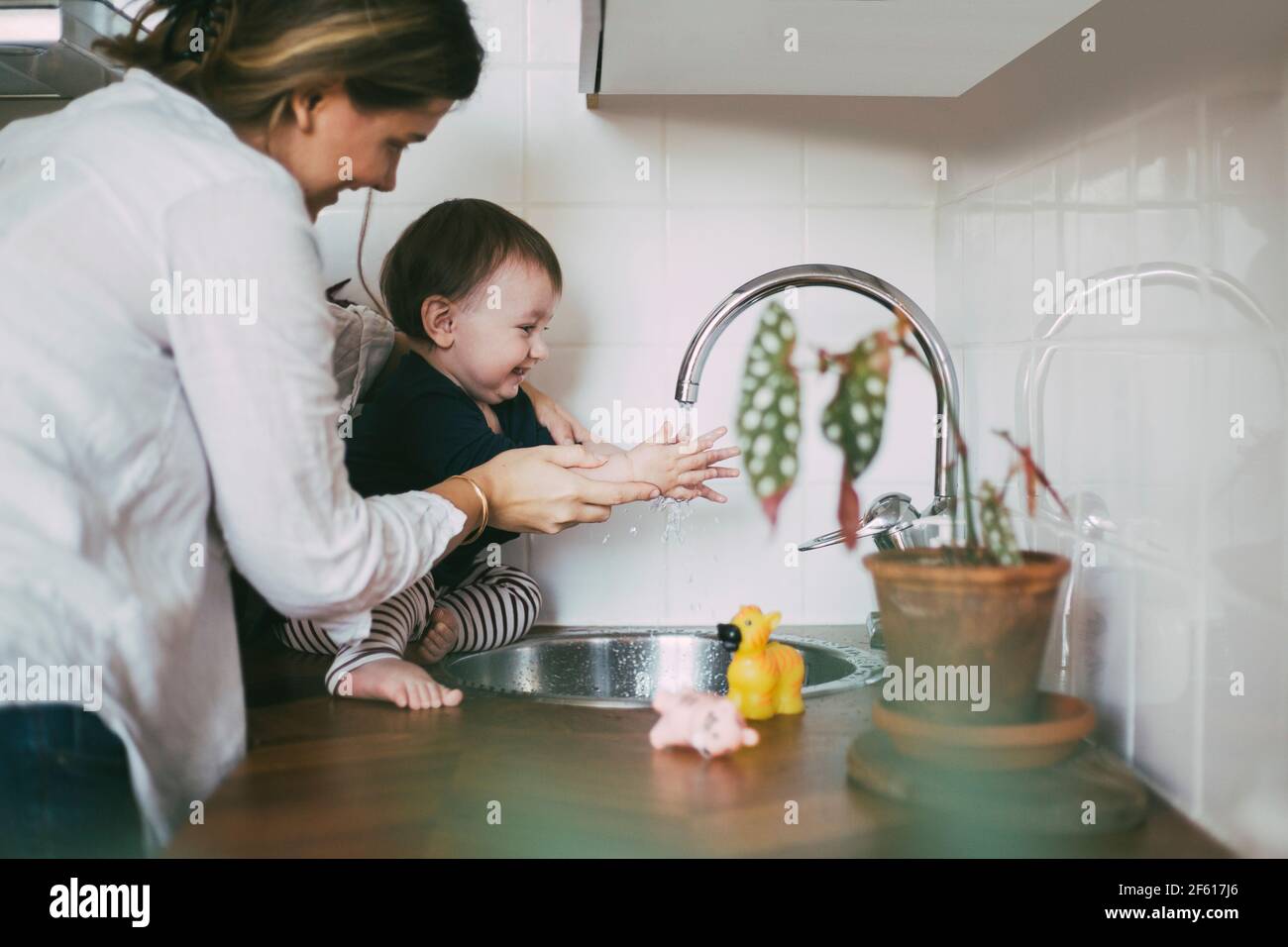 Smiling mother helping baby girl washing hands under sink in kitchen ...