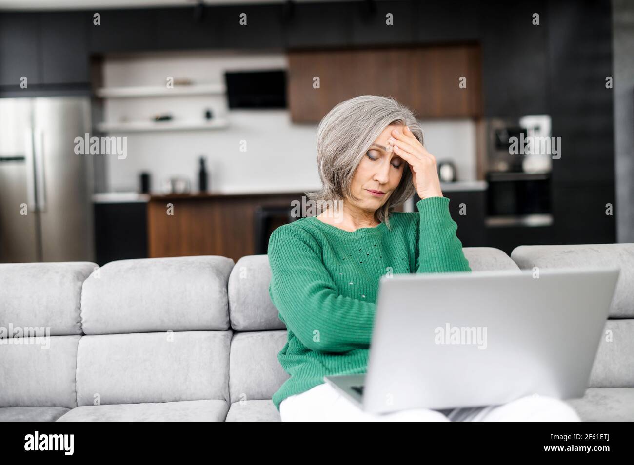 Upset senior woman sits with a laptop on the couch at home, sad because ...