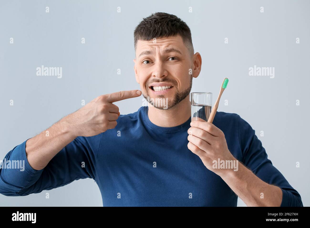 Man with aching teeth, brush and glass of water on grey background ...