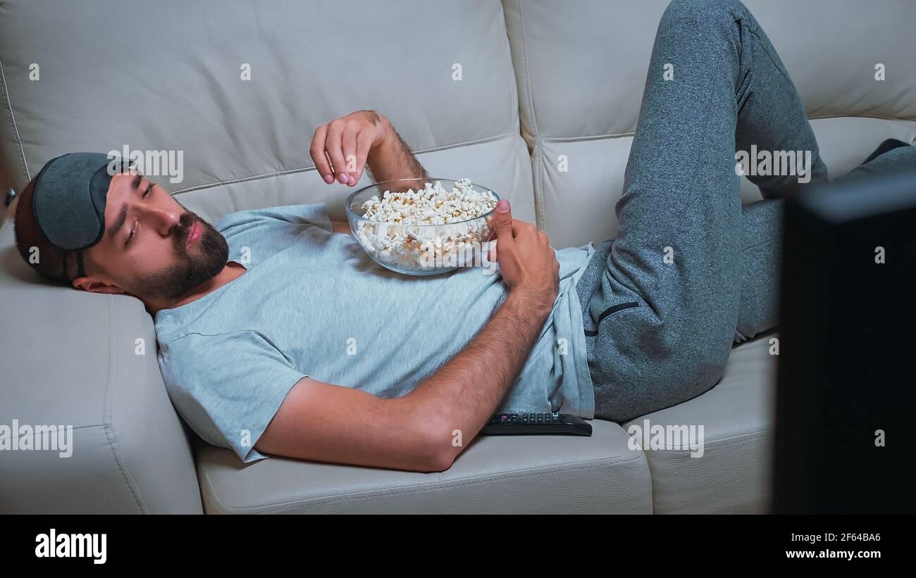 Tired man relaxing on sofa in front of television eating popcorn while ...
