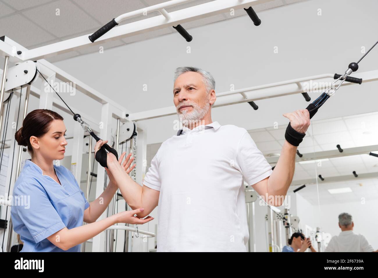 brunette rehabilitologist assisting middle aged man working out in ...