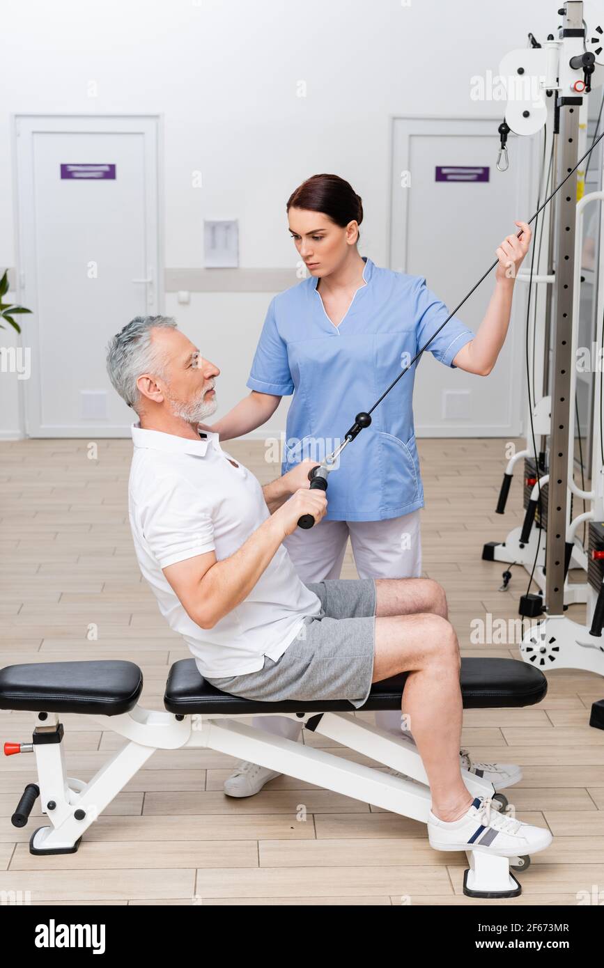 hospital trainer assisting middle aged man working out on pull cable ...