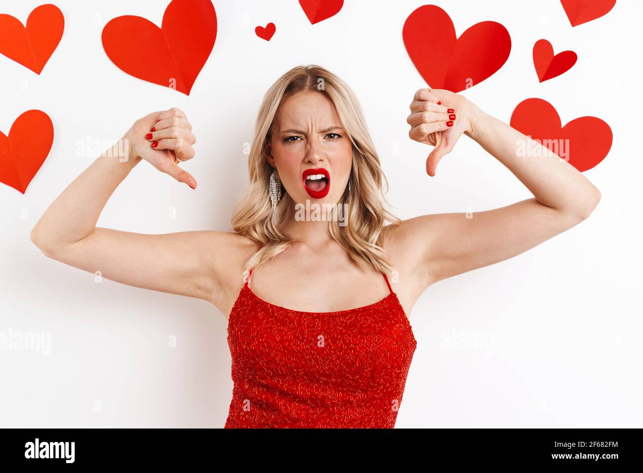 Upset blonde girl in red dress gesturing dislike sign with thumbs down ...