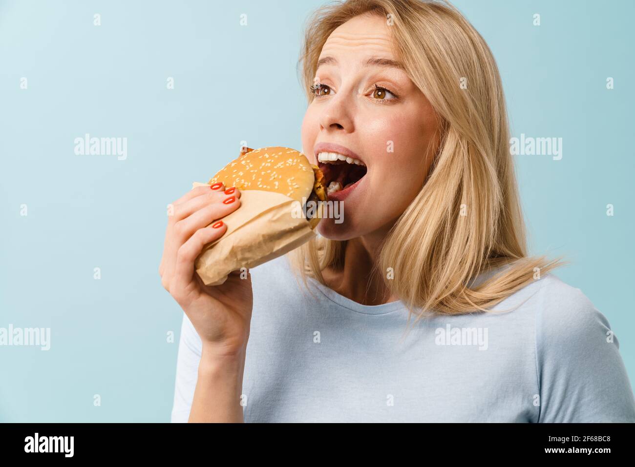 Cheerful hungry blonde girl eating hamburger on camera isolated over ...