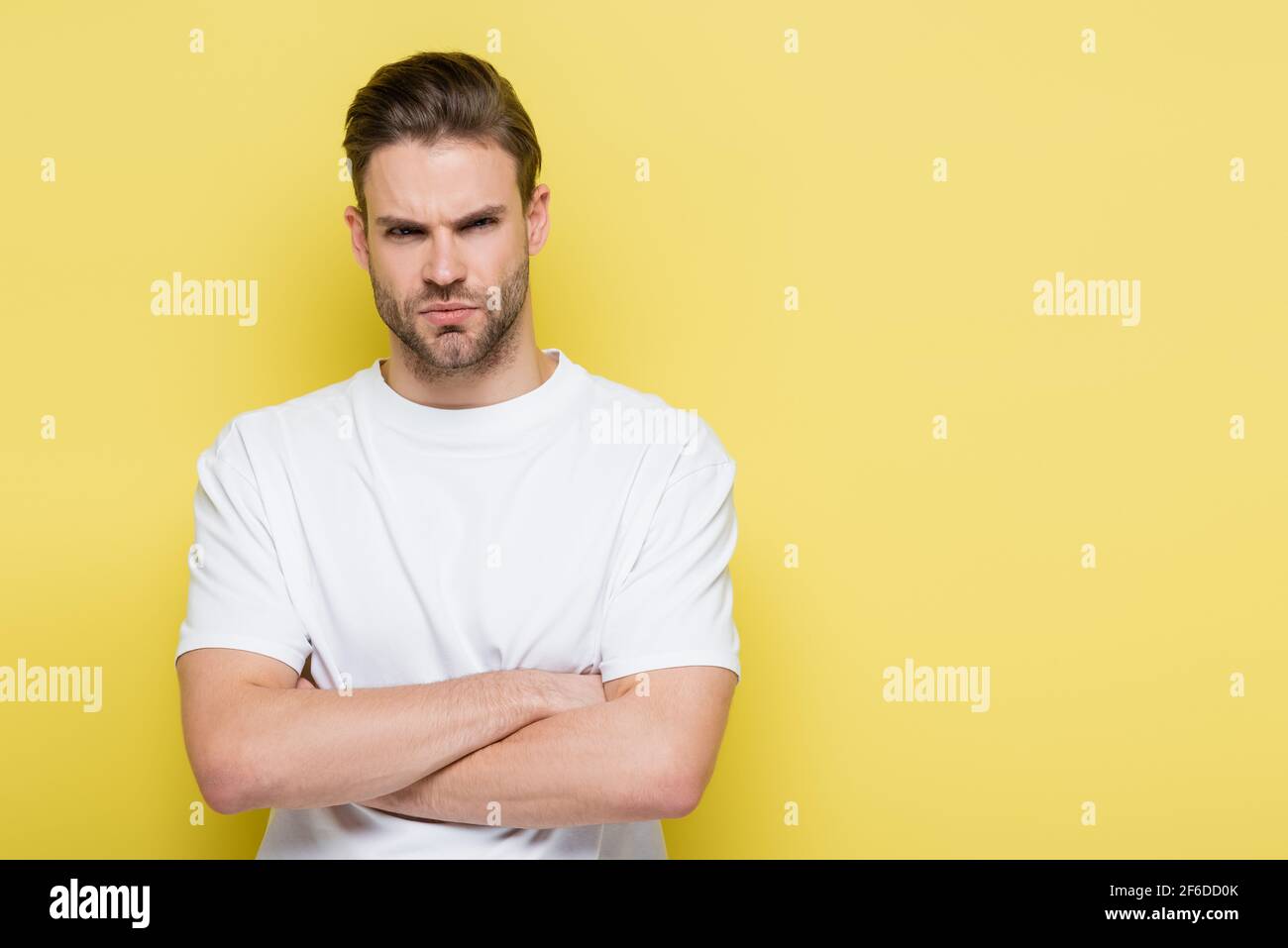 angry man looking at camera while standing with crossed arms on yellow ...