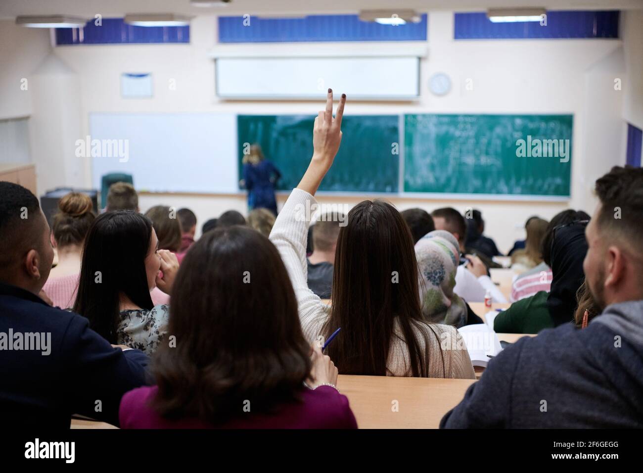 Rear view of female student sitting in the class and raising hand up to ...
